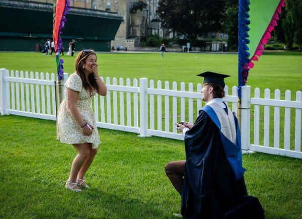 A graduate in ceremonial cap and gown, bends down on one knee in front ...